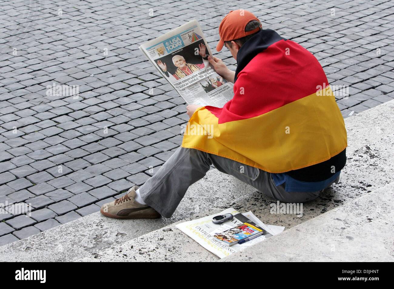 (dpa) - A German pilgrim covered by a German flag reads a newspaper ...