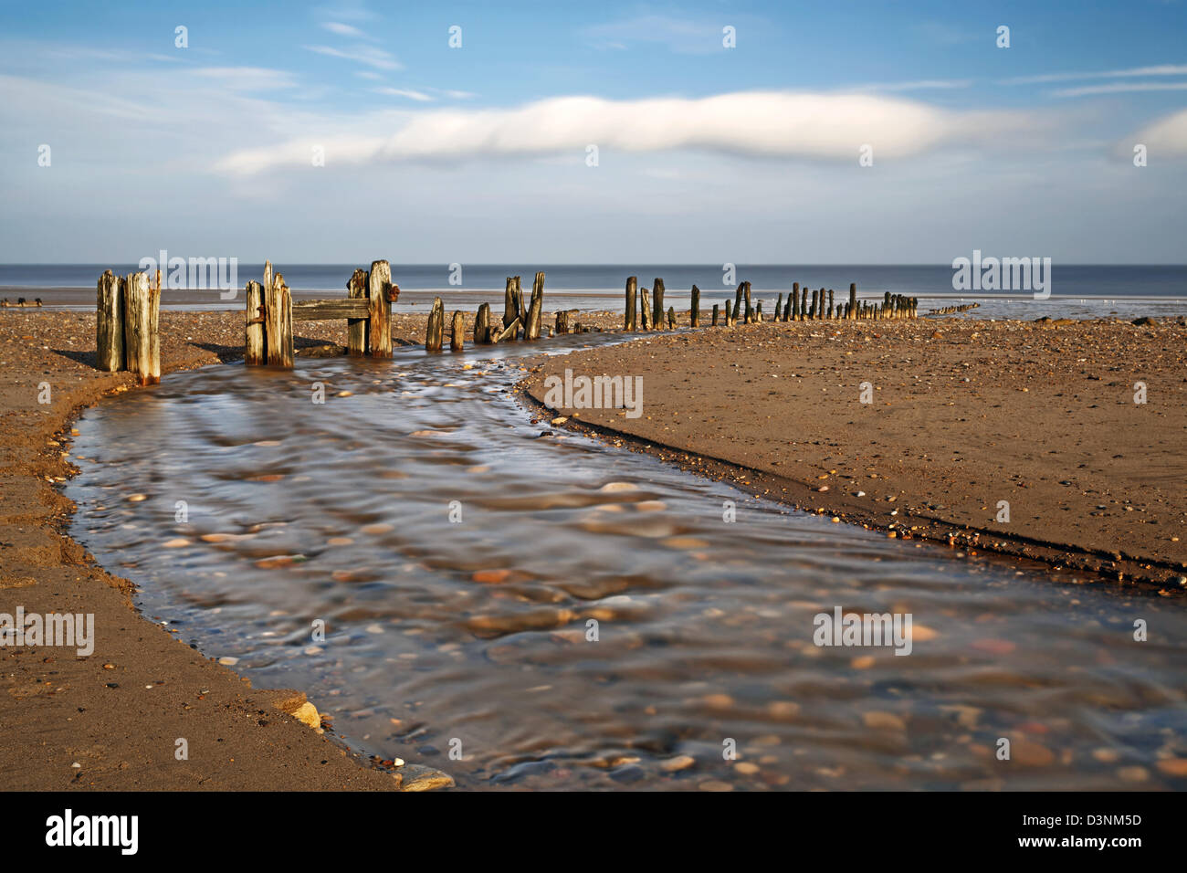 A small estuary flowing through the sand besides old wooden groynes at ...
