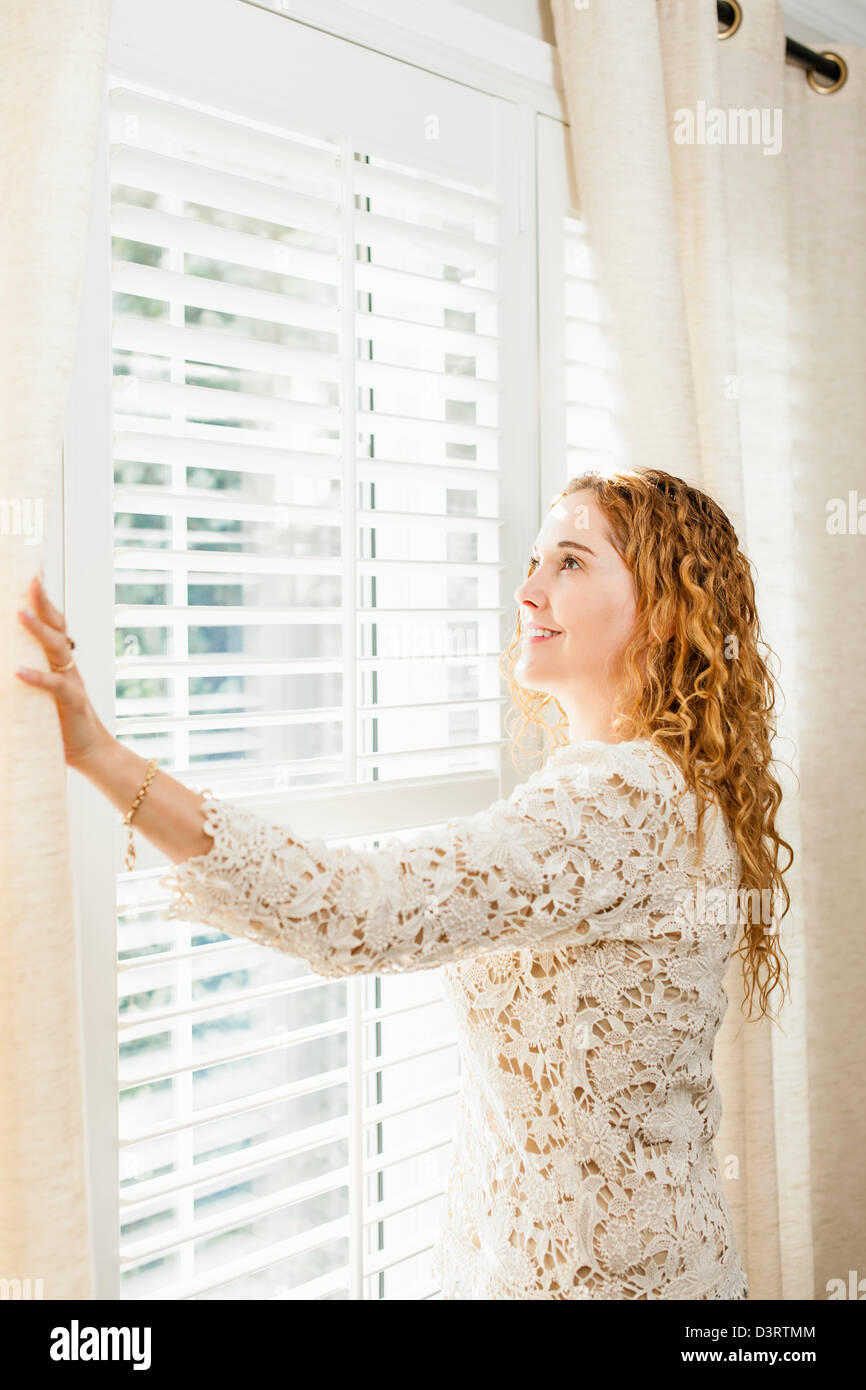 Happy woman looking out big bright window with curtains and blinds ...