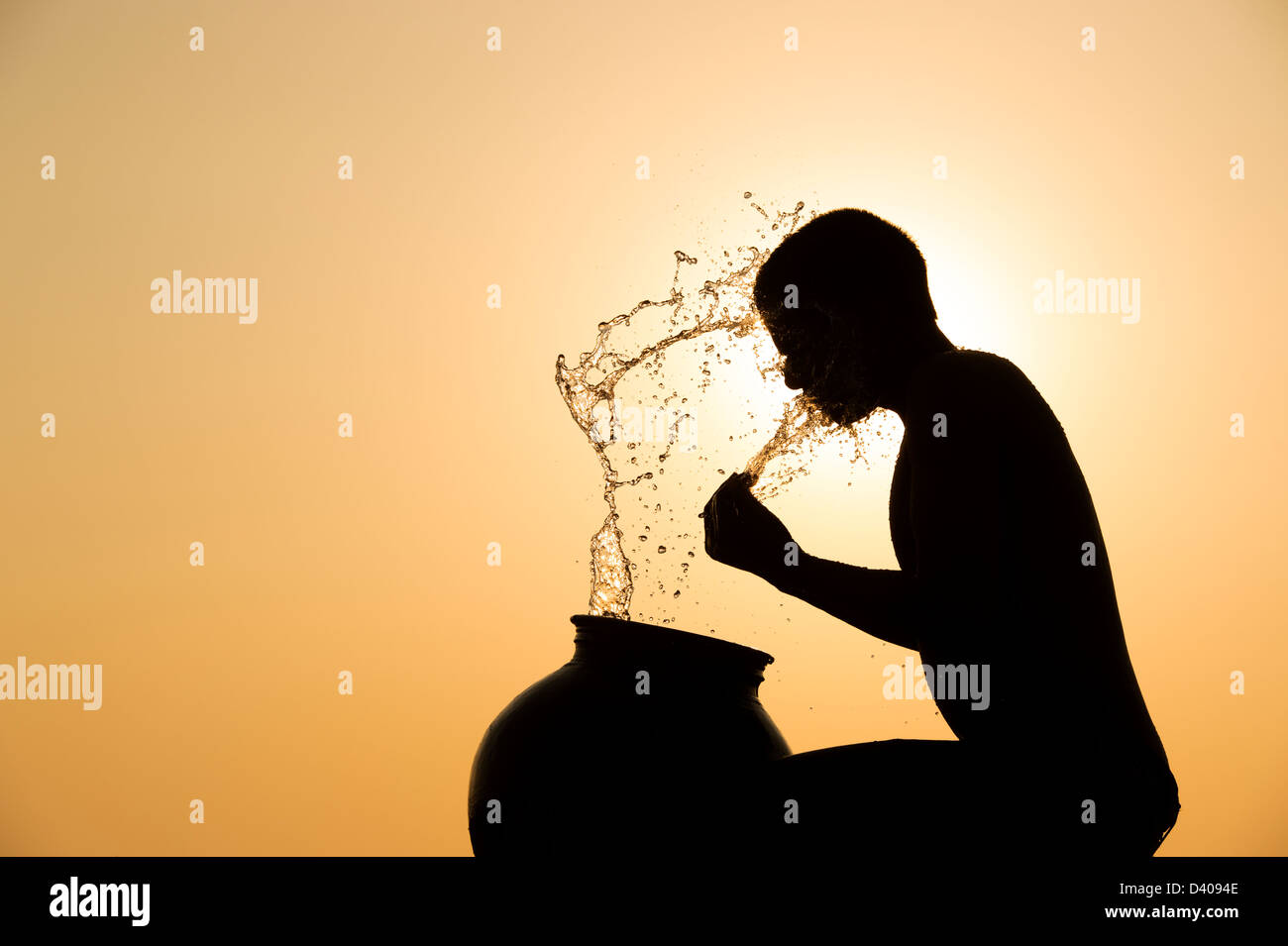Indian man splashing water over himself from a water pot at sunset ...