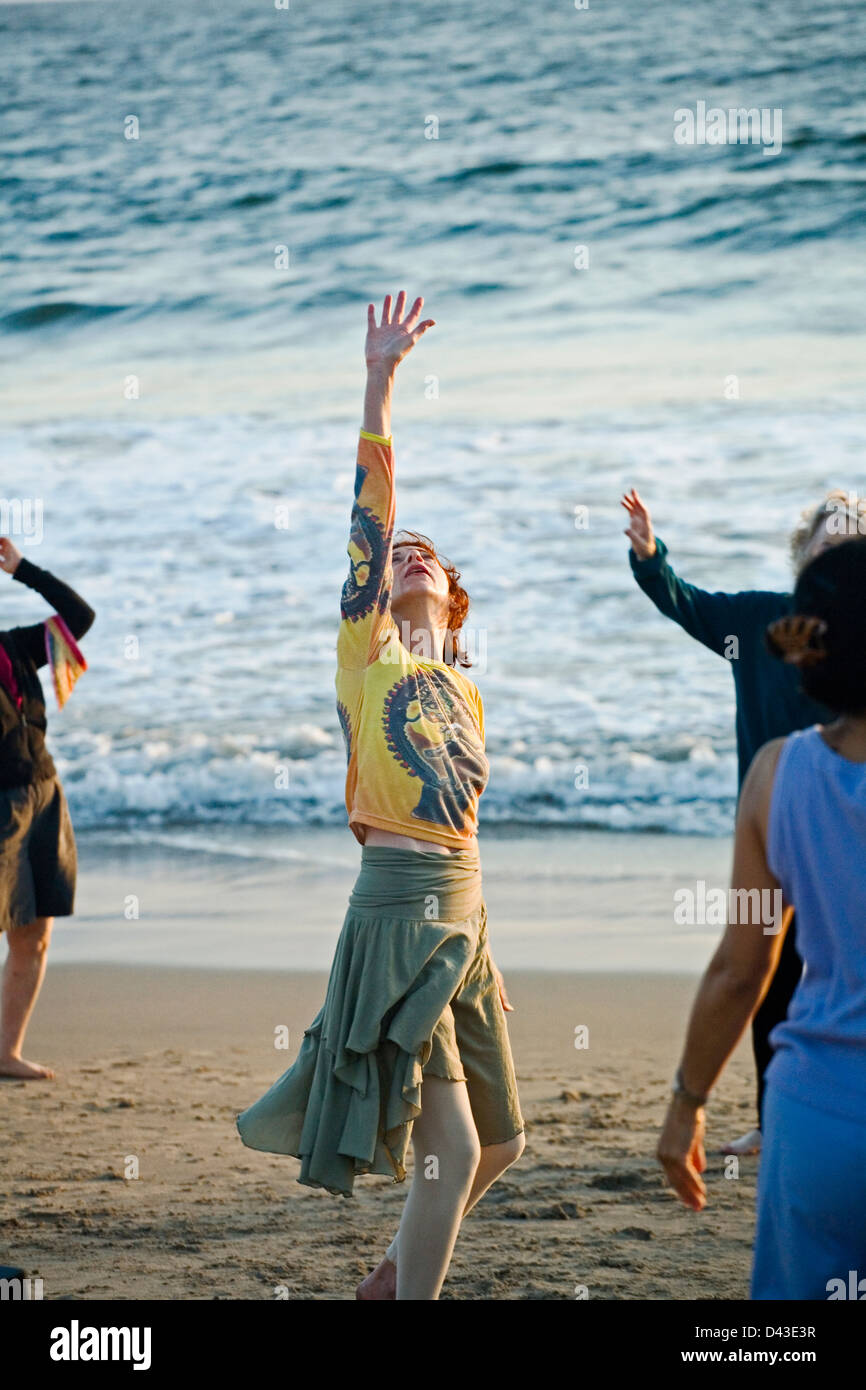 Woman reaches up to sky with ocean behind her as she dances and ...