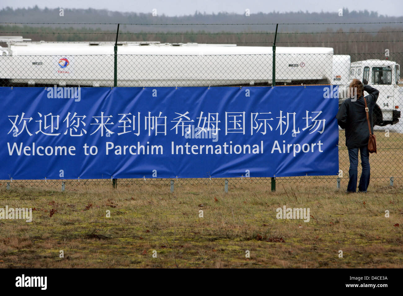 A woman stands in front of a fence with a banner 'Welcome to Parchim ...
