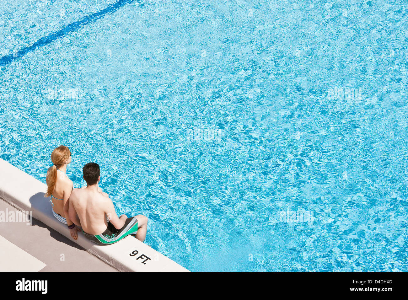 A young couple wearing bathing suits, sitting on the edge of a swimming ...