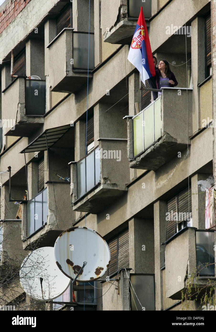 Two people fly a Serbian flag on a balcony with rotten satellite dishes ...