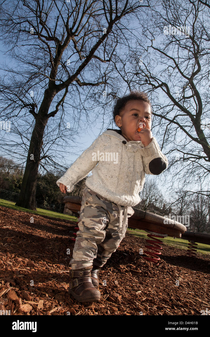 mixed race toddler boy in park eating orange walking with trees behind ...