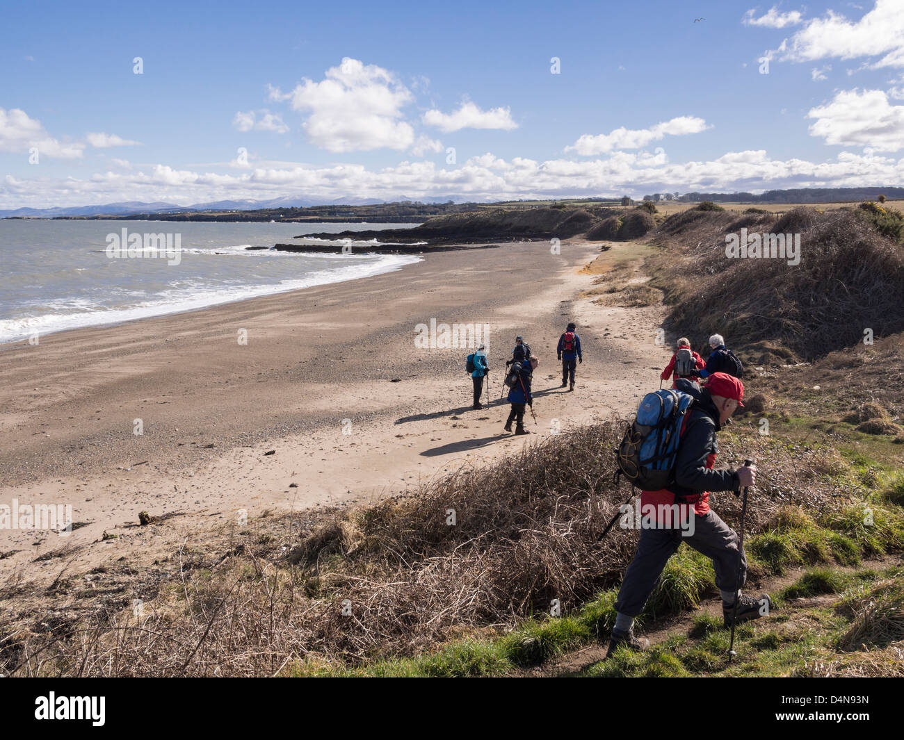 Ramblers walking on the coastal path across quiet Traeth Lligwy beach ...