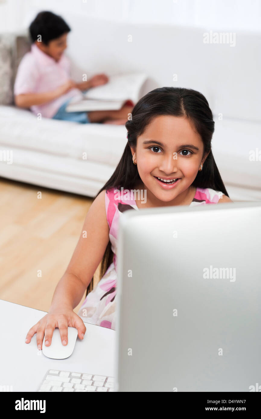 Girl using a computer with her brother reading a book in the background ...
