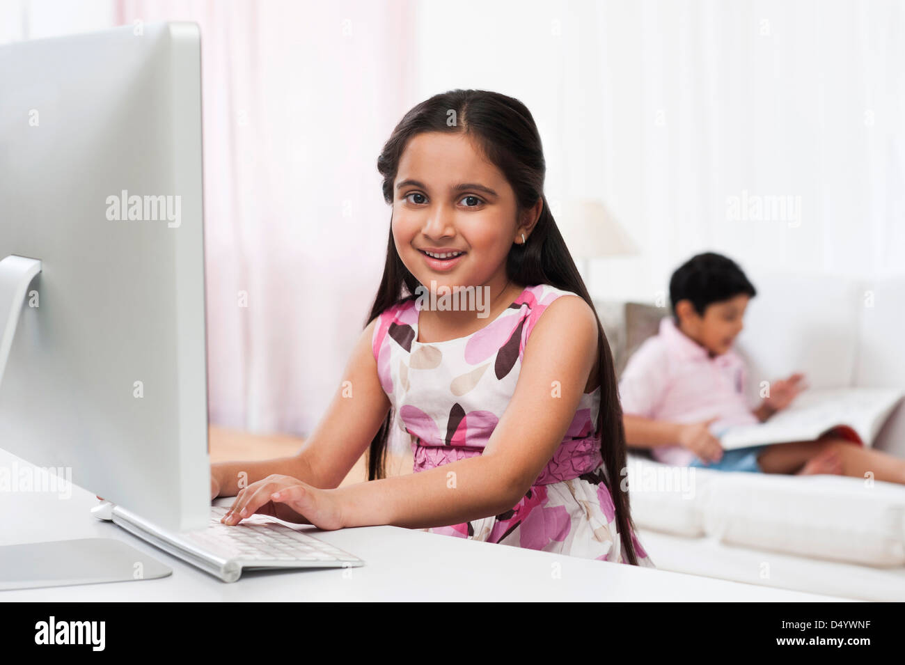 Girl using a computer with her brother reading a book in the background ...
