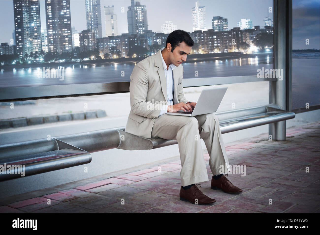 Businessman Sitting On A Bench At Bus Stop And Working On A Laptop