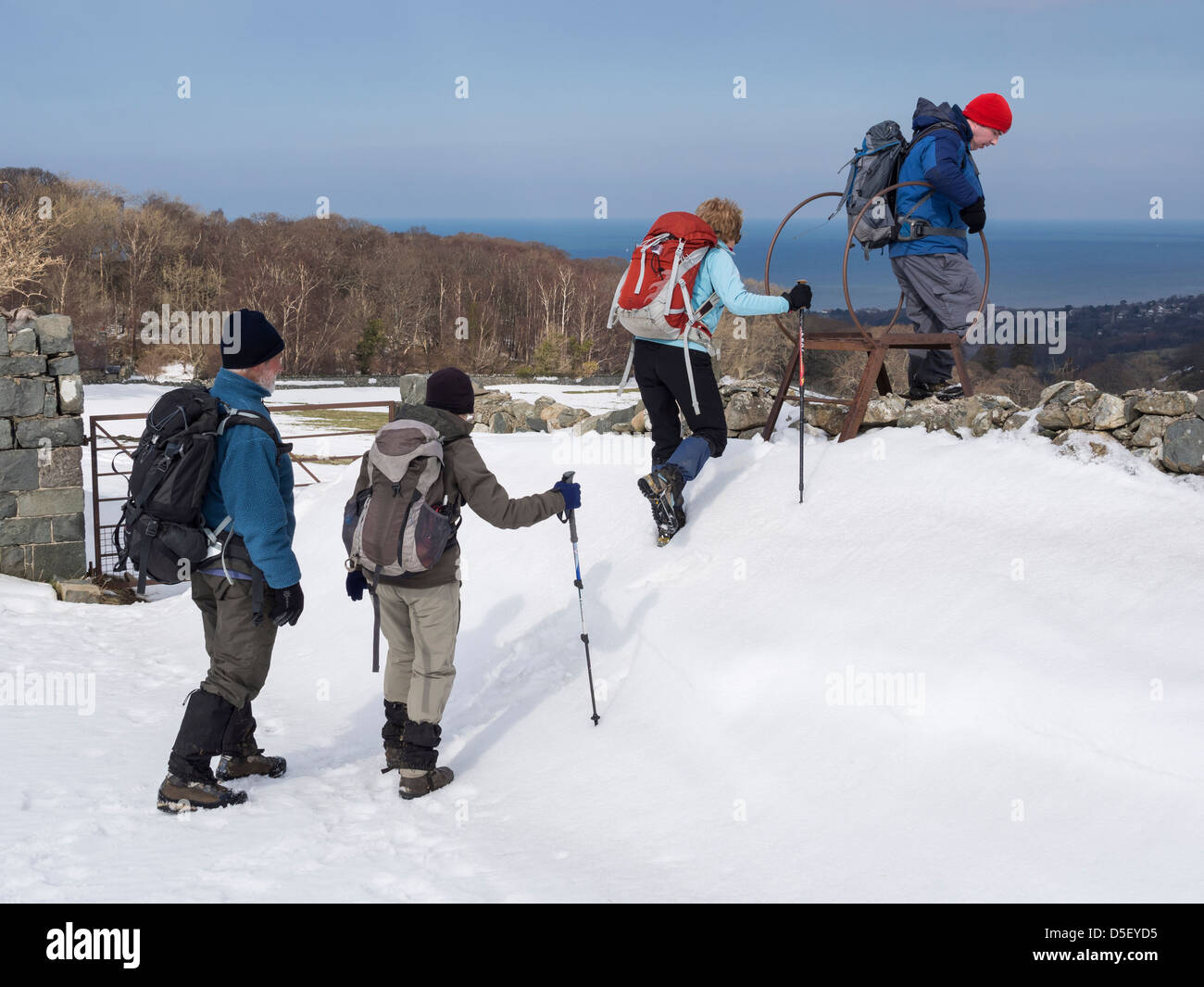 Ramblers with rucksacks walking up a deep snow drift by ladder stile ...