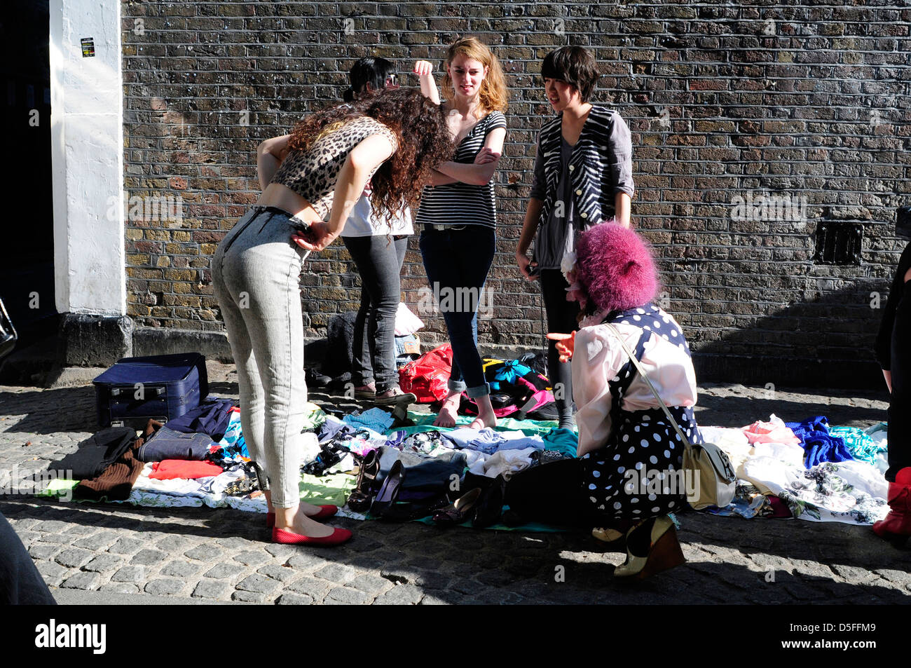 A group of young women selling second hand clothes on a pavement. Brick ...