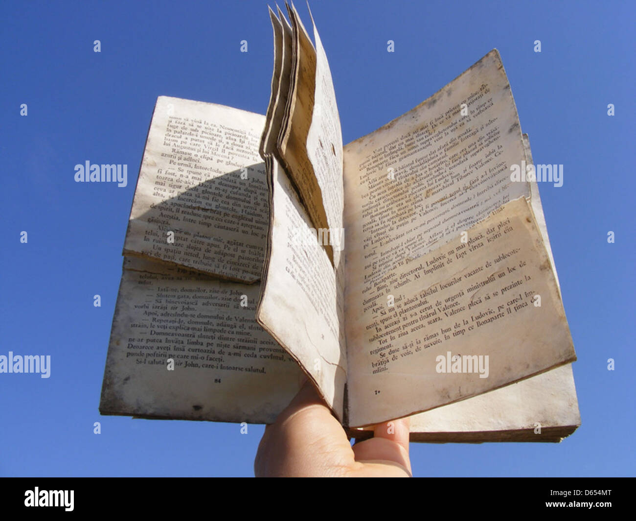 A hand holds a dusty, old book under a clear blue sky. The image ...
