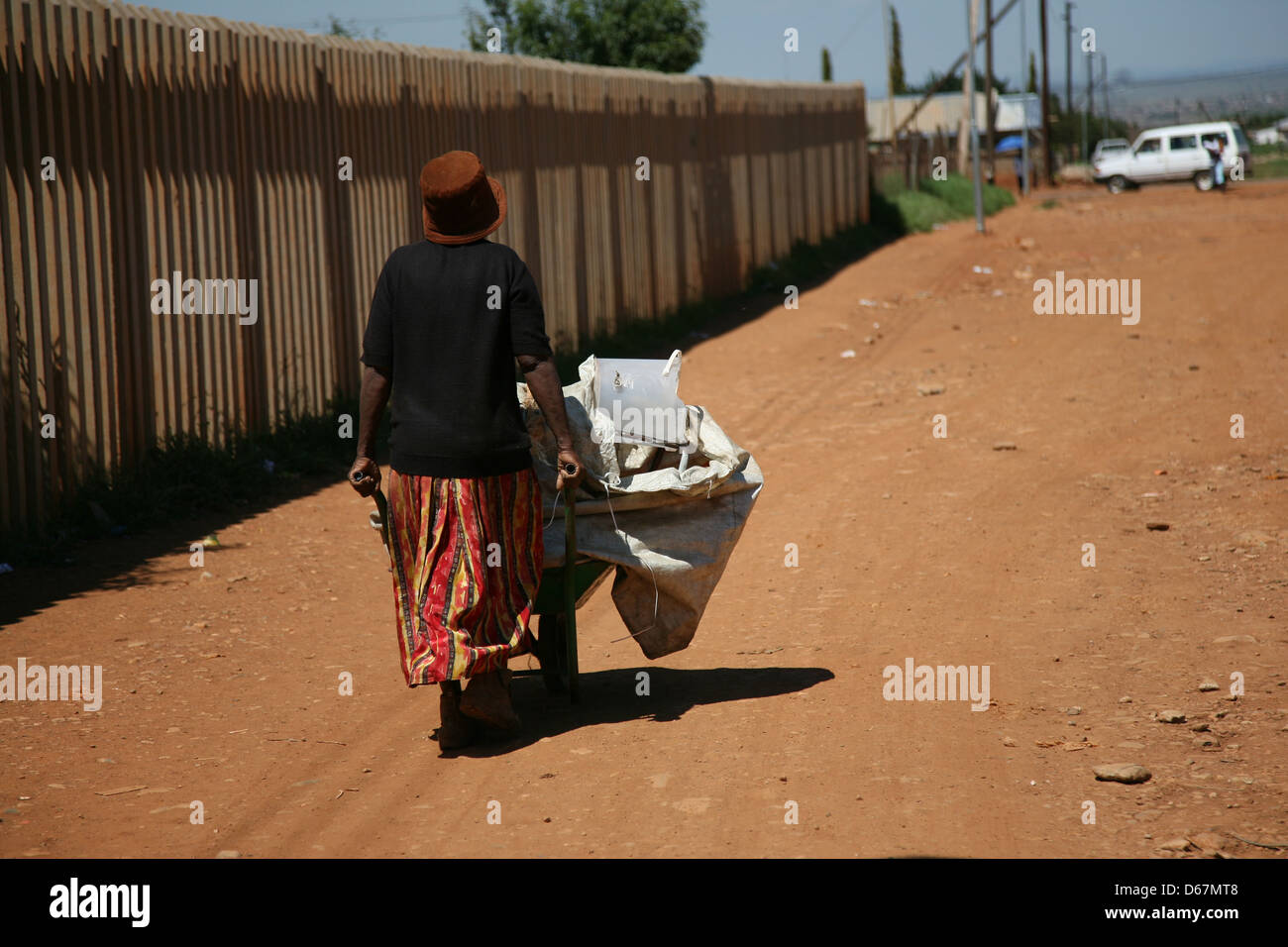 An African woman tansporting goods on a wheelbarrow in a rural housing ...