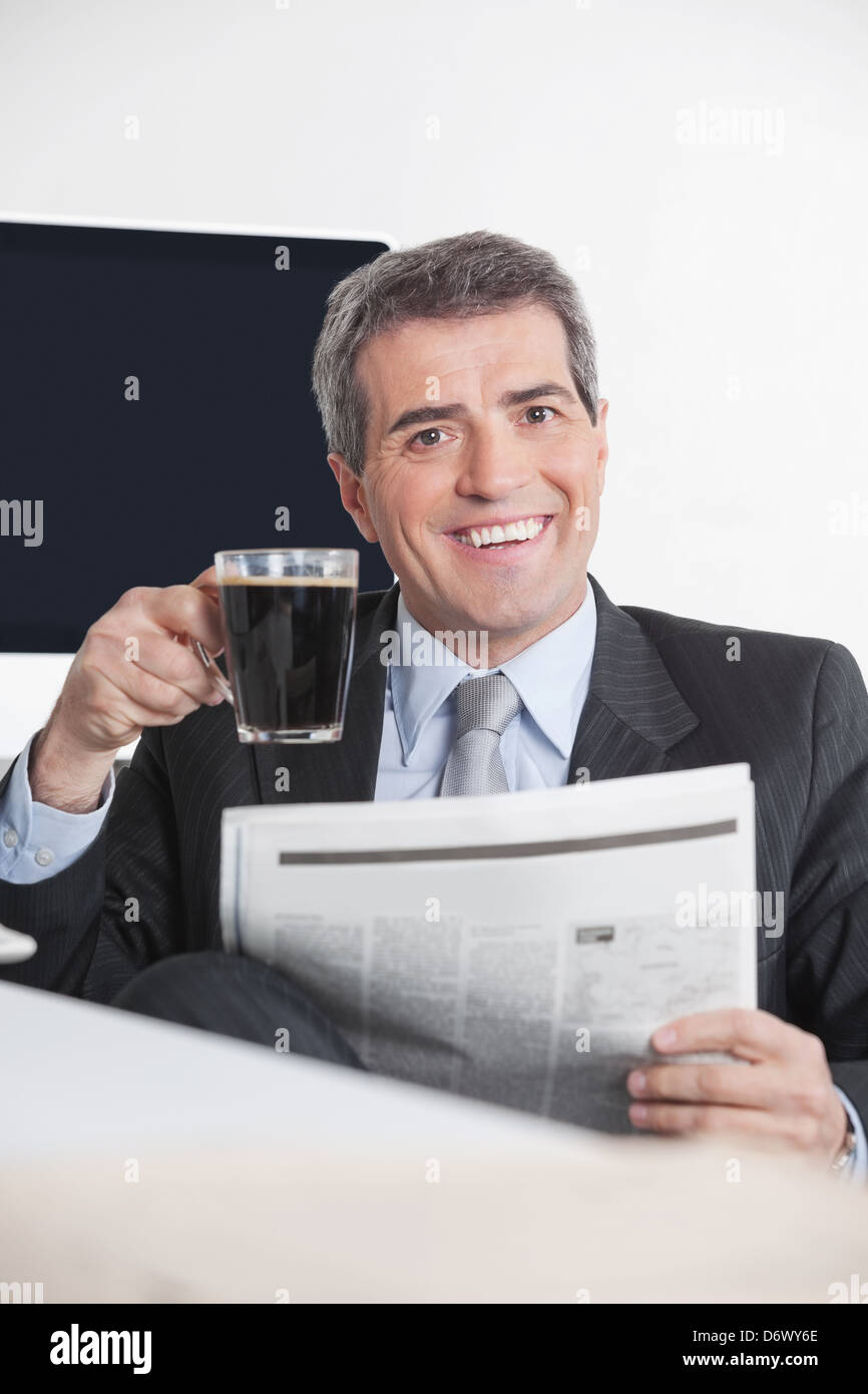 Happy manager sitting with newspaper and coffee at his desk in the ...