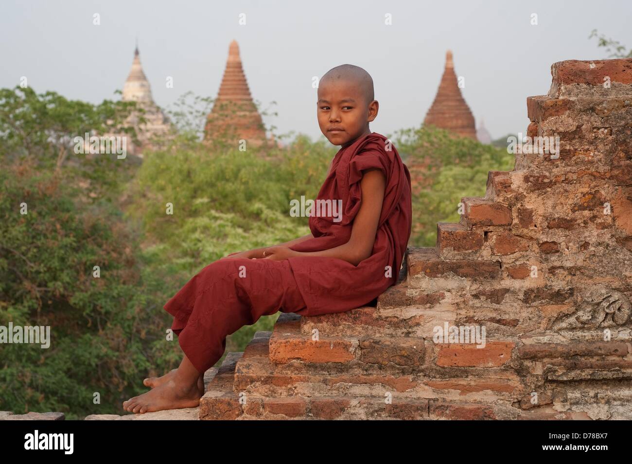 A young Buddhist monk is sitting on a brick wall in front of a Pagoda ...