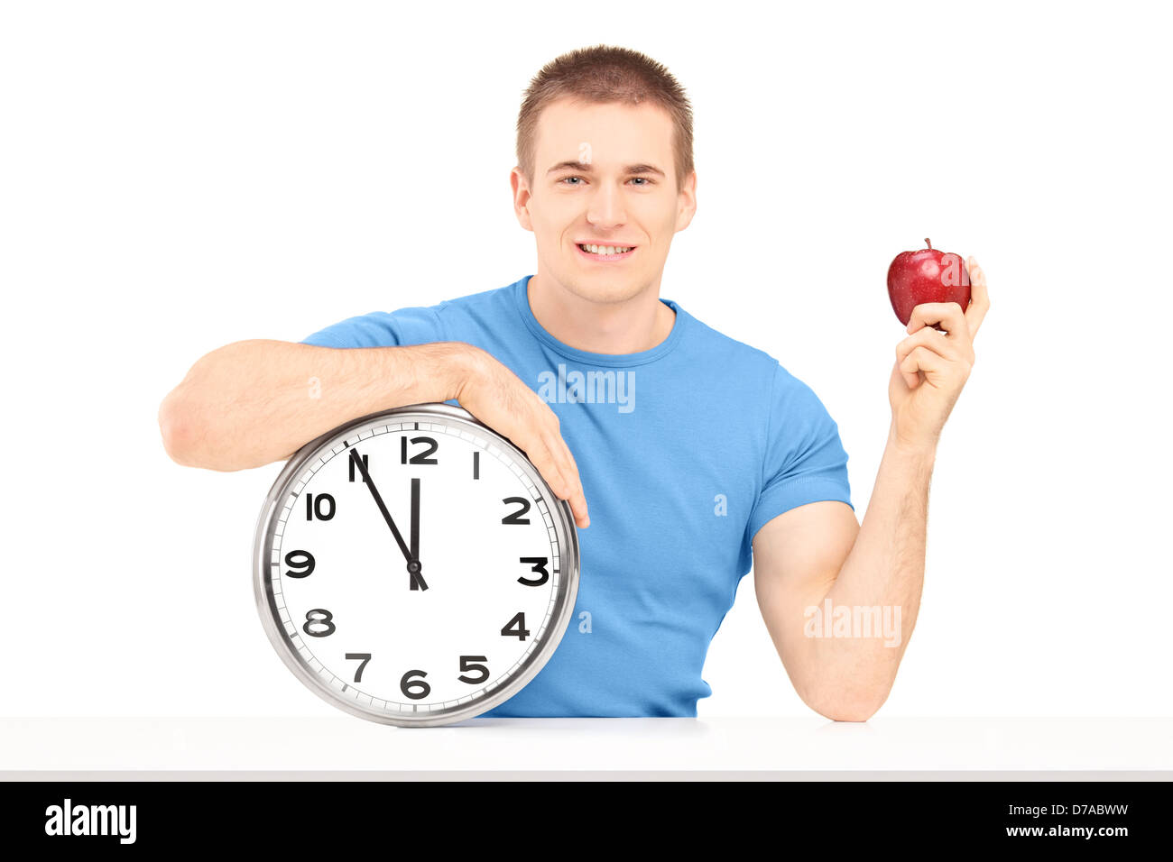 A smiling guy holding a wall clock and red apple on a table isolated on ...