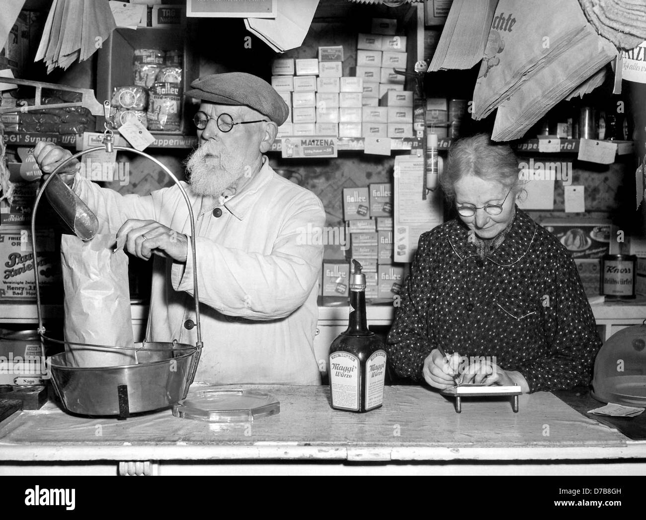 Two elderly people work in their grocer's shop between shelves full of ...