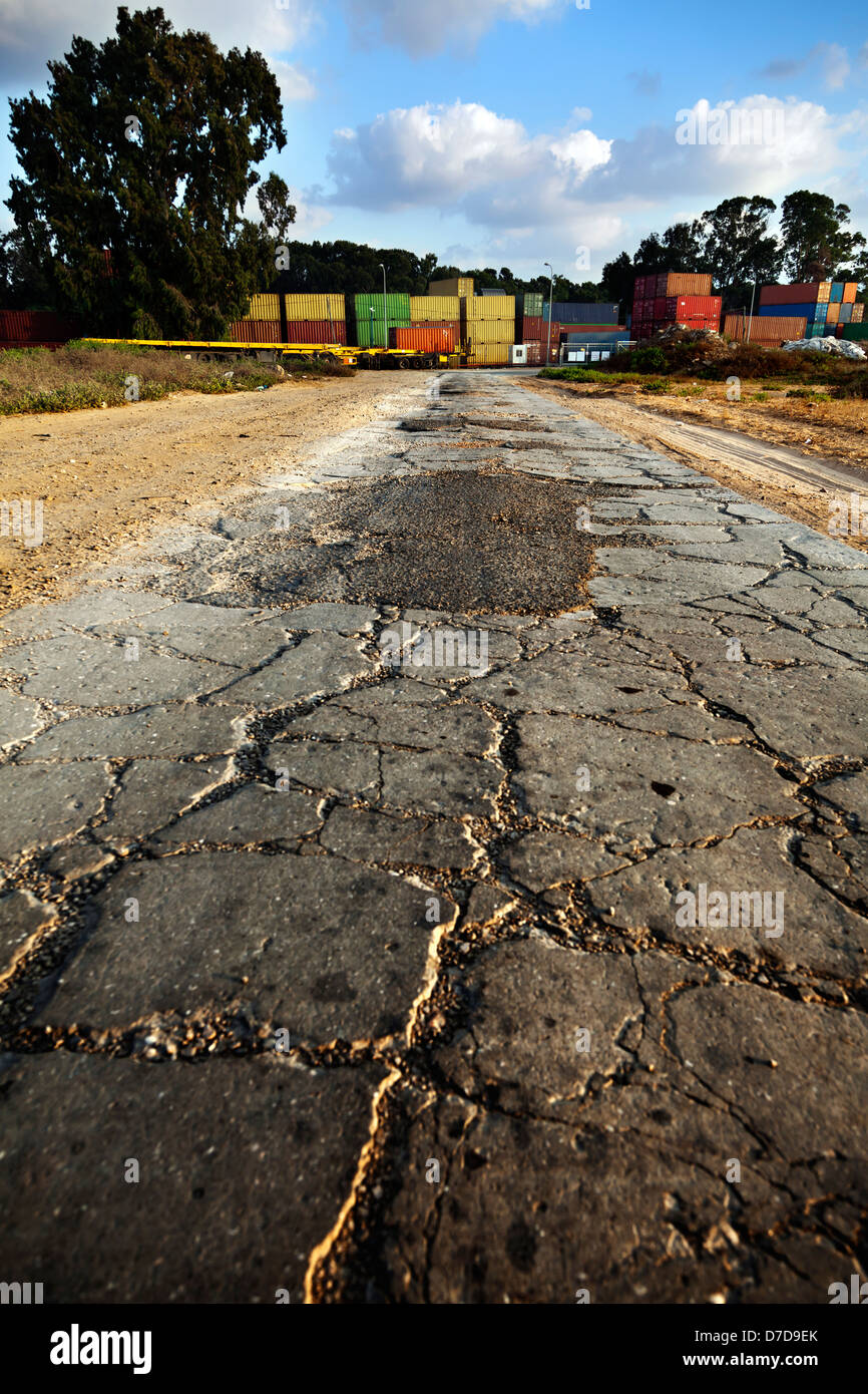 A badly patched broken asphalt road leads to a storage yard of cargo ...