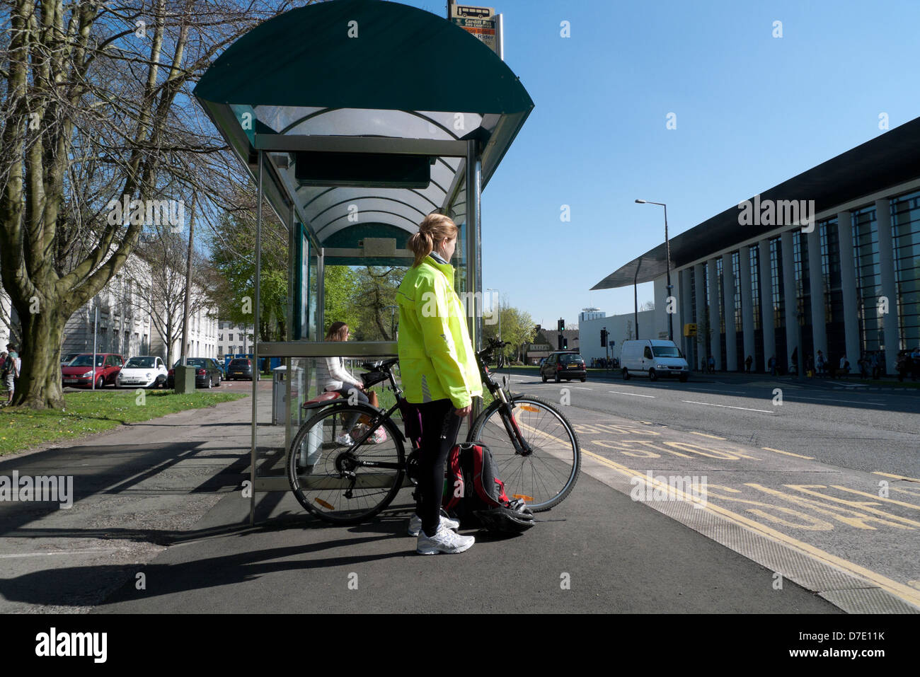 Female cyclist with a fluorescent jacket waiting at a bus stop across ...