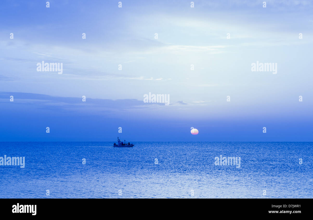 A small fishing boat in a boundless sea advances slowly in the light of ...