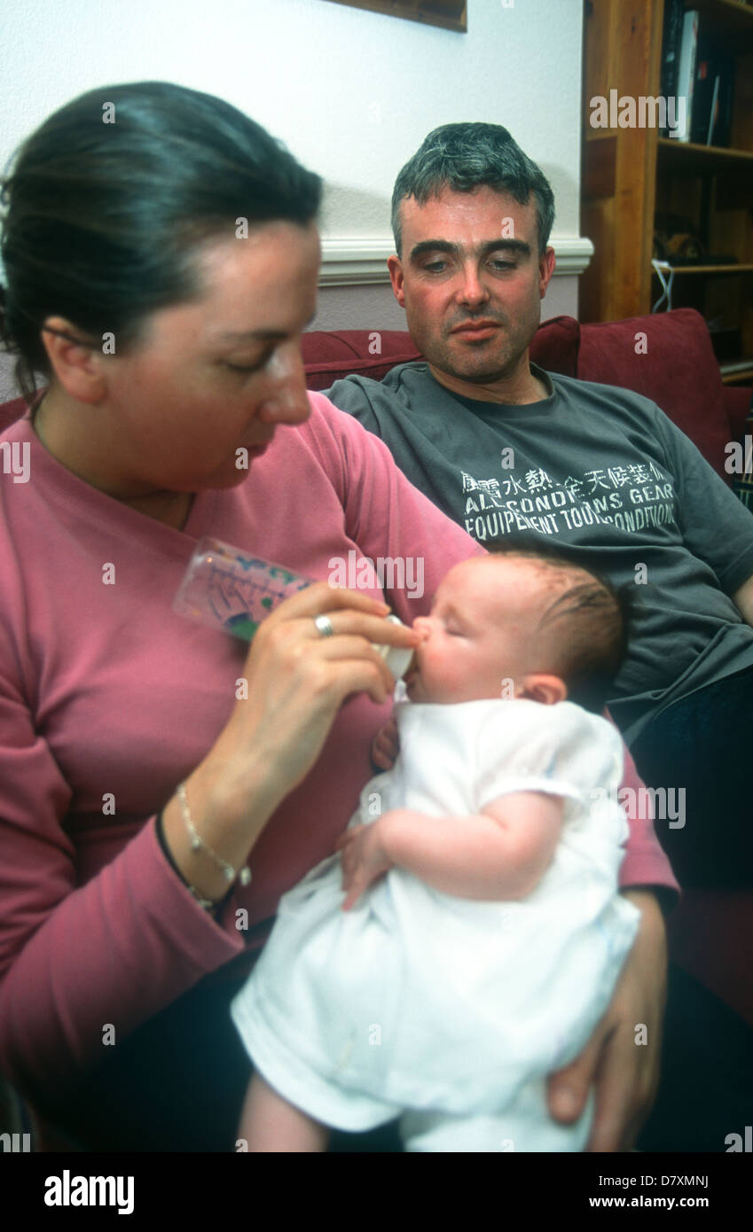Irish mother bottle feeding baby girl whilst dad looks on in background ...