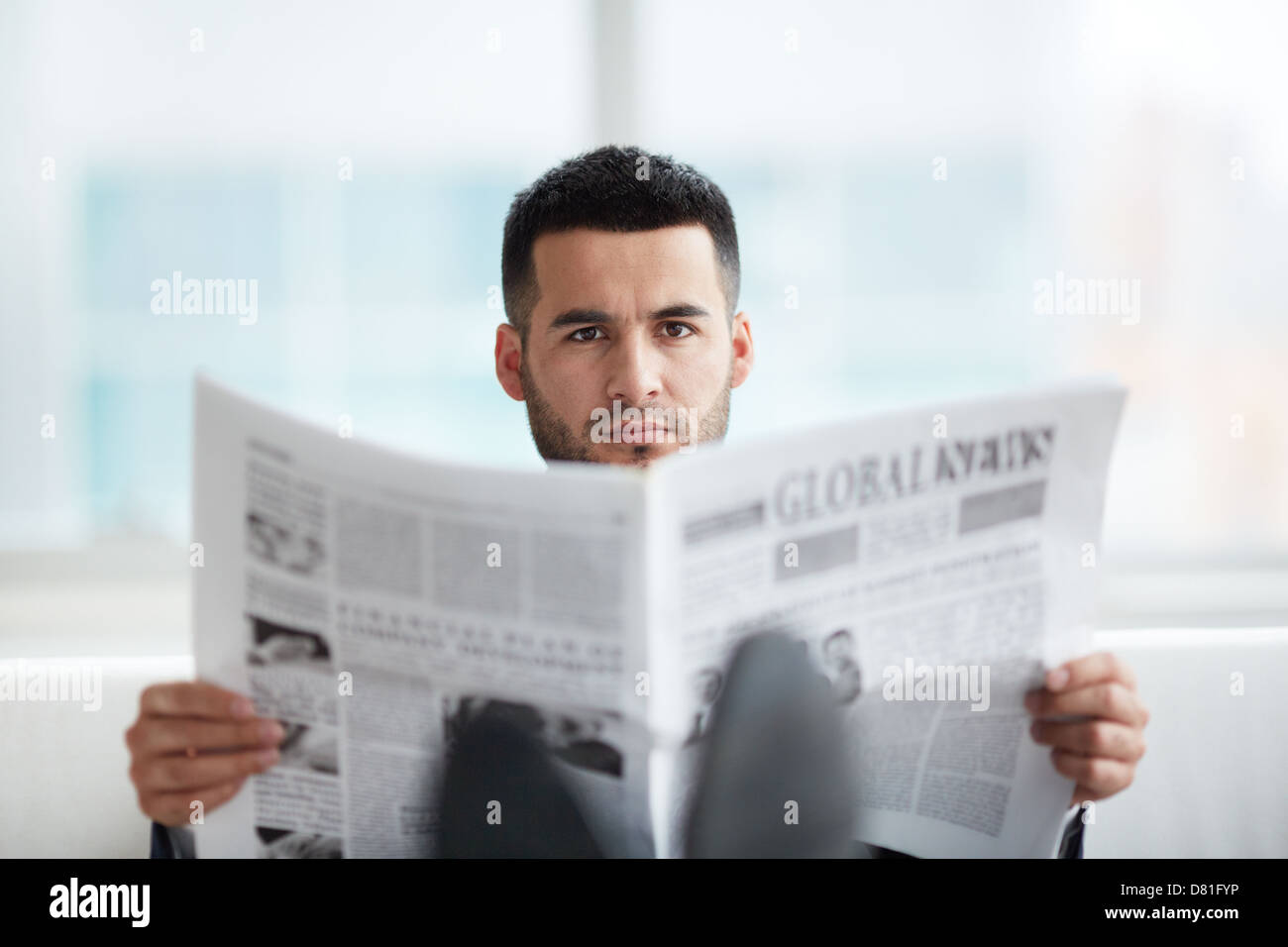 A young serious businessman looking at camera while reading newspaper ...