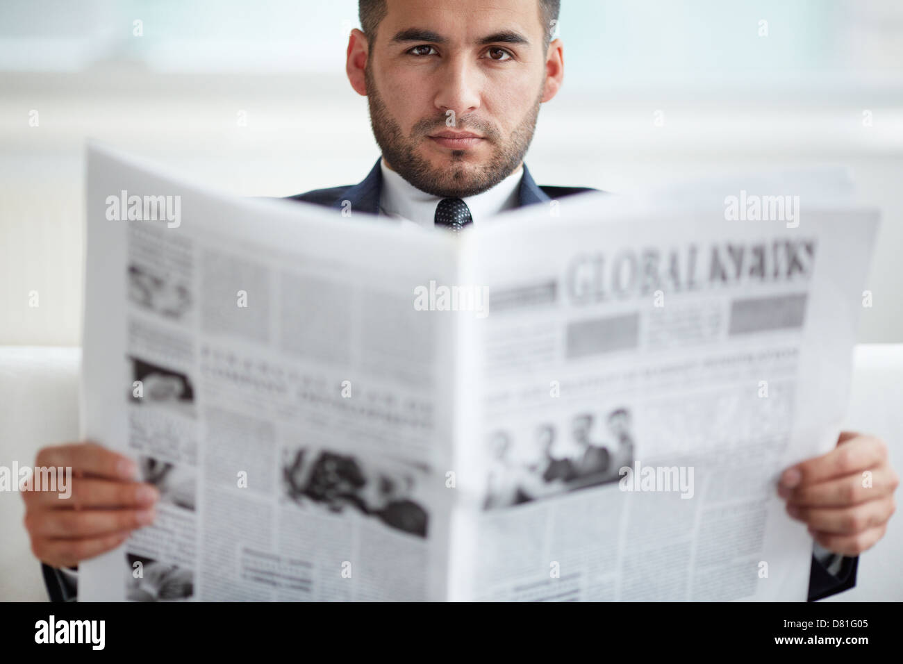 A young serious businessman looking at camera while reading newspaper ...