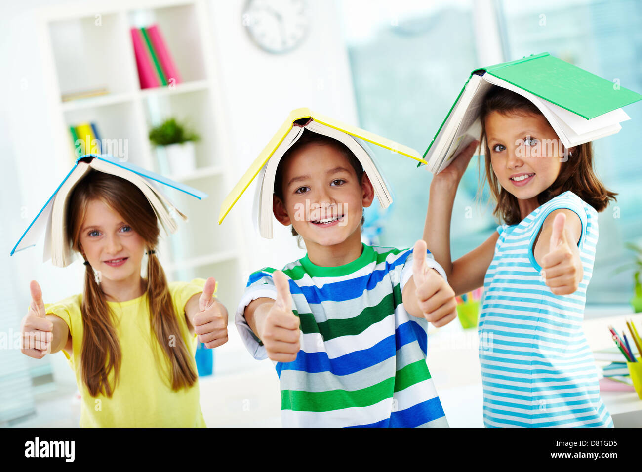 Portrait of happy classmates with books on their heads keeping thumbs ...