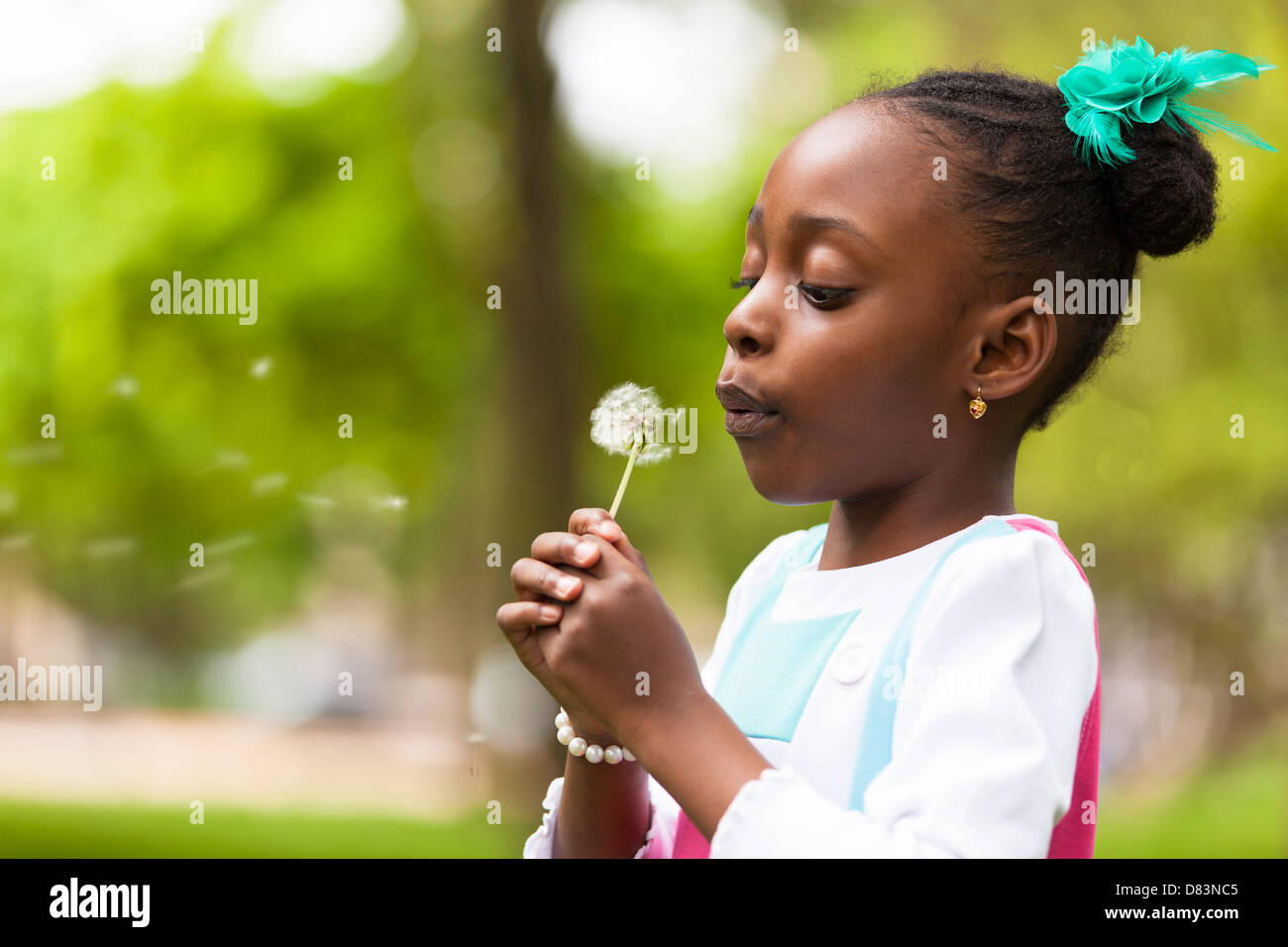 Outdoor portrait of a cute young black girl blowing a dandelion flower ...