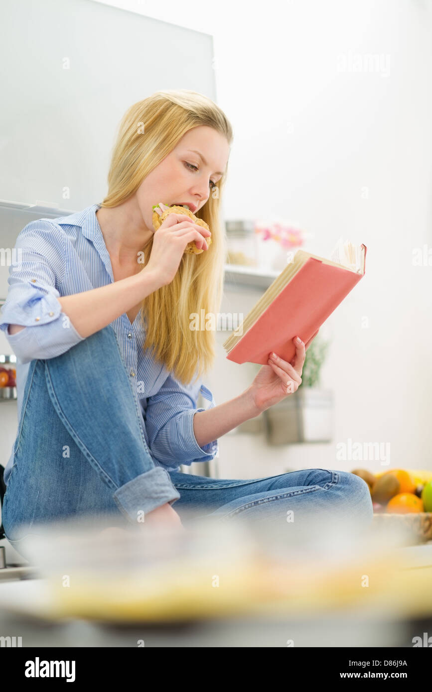 Young woman eating sandwich and reading book in kitchen Stock Photo - Alamy