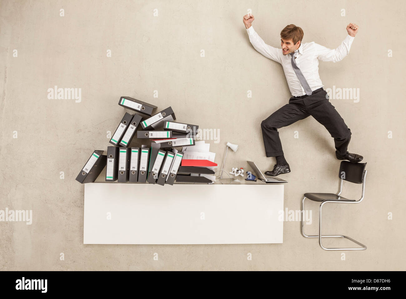 Businessman jumping from office desk and chair besides stack of files ...