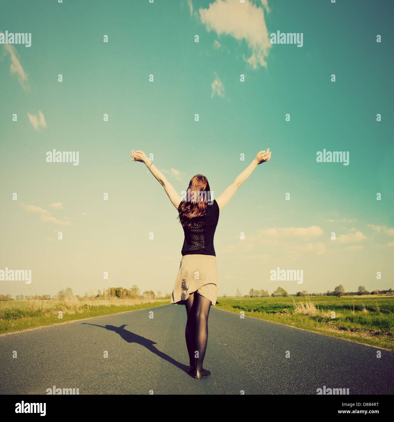Happy woman standing with hands up on long straight road facing the sun ...