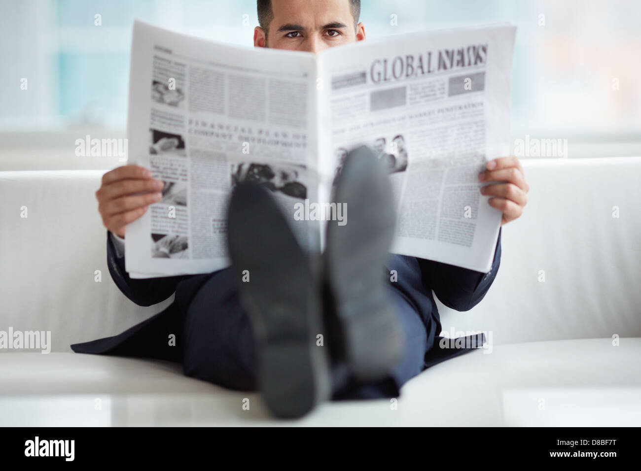 A young businessman on sofa looking at camera while reading newspaper ...