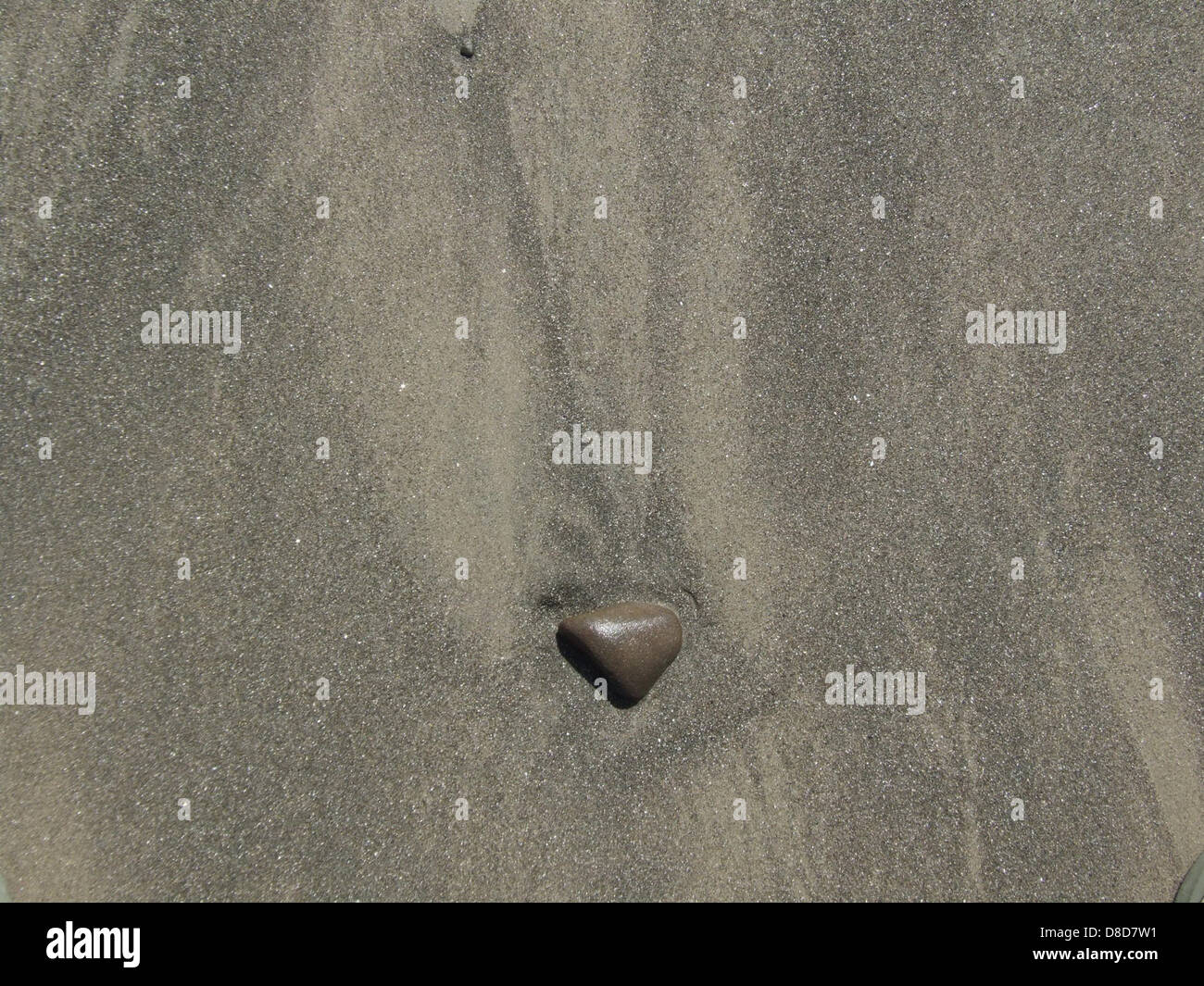 A scene of sand on a beach, with a large stone partially buried in the ...