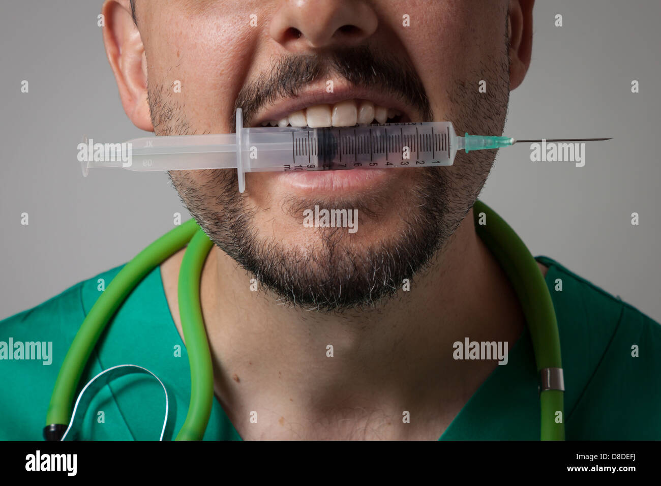 Funny young physician in green uniform holding a syringe with his teeth ...
