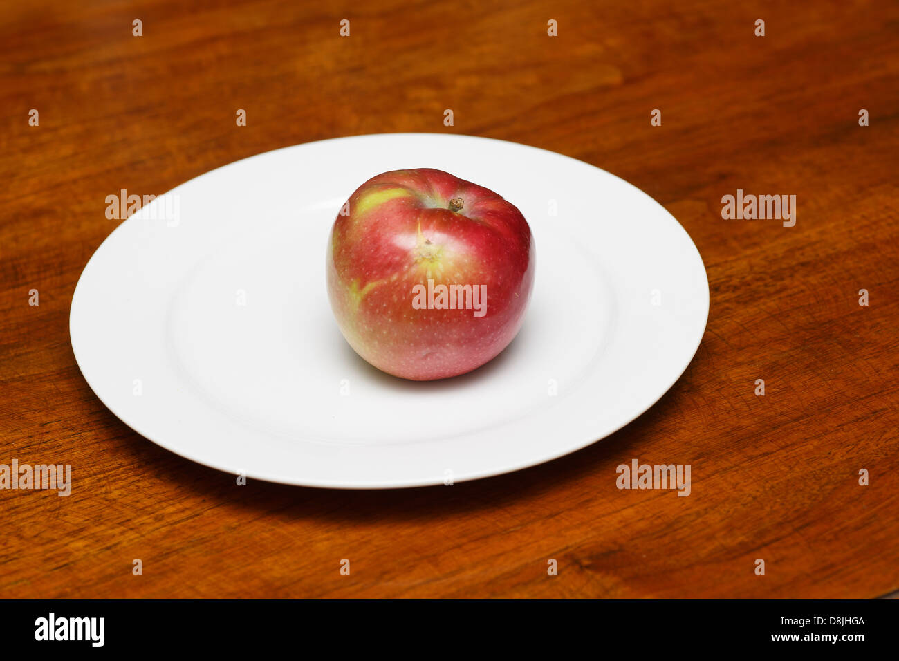 A single red apple alone on an empty white plate and wood table Stock ...