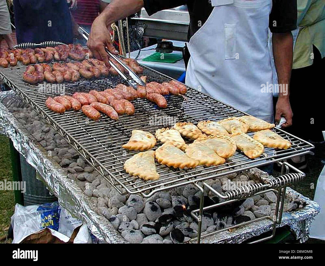 This image shows a barbecue scene with sausages and empanadas being ...