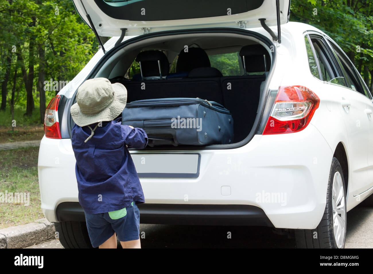 Small boy loading his suitcase into the open back of a hatchback car as ...