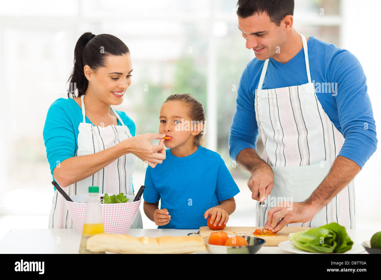 little girl tasting tomato while her parents cooking in kitchen at home ...