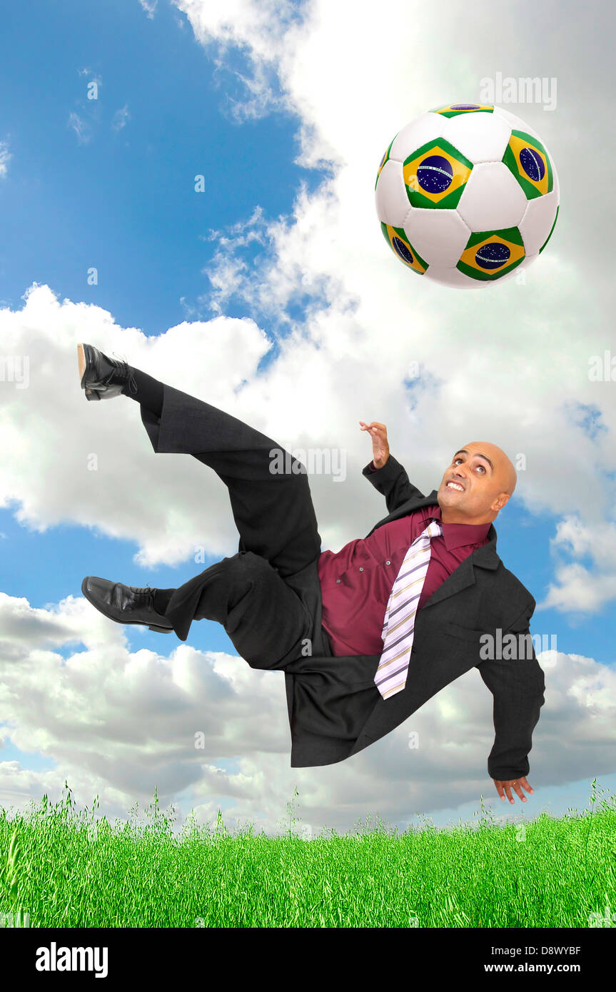 Businessman in a acrobatic pose kicking a soccer ball in a green field ...