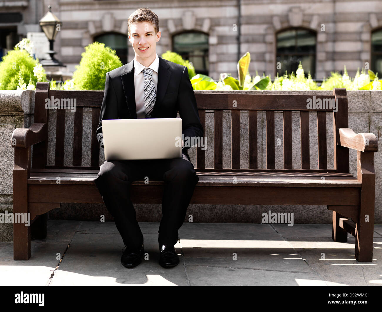 A cheerful young businessman using a laptop while sitting outdoors on a ...