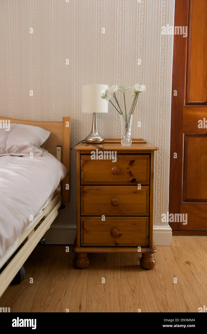 A set of bedside drawers at the side of a bed with some flowers and a ...