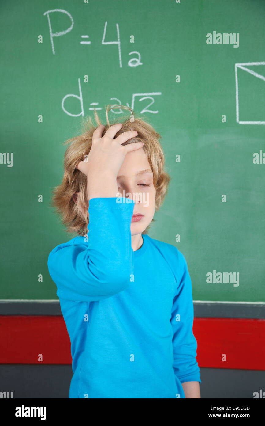 Sad little schoolboy standing with hand in hair by board in classroom ...