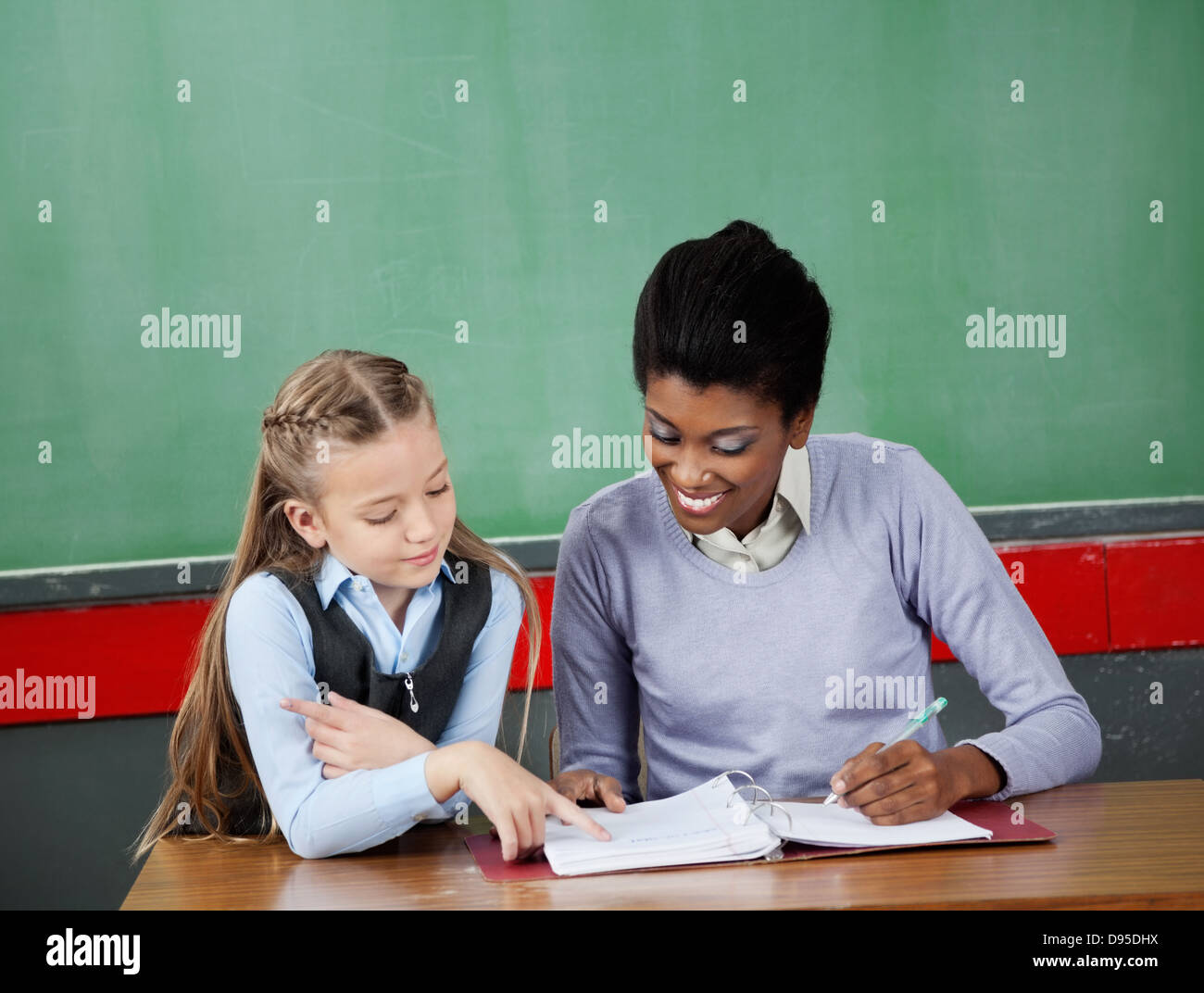 Little schoolgirl asking question to female teacher at desk in ...