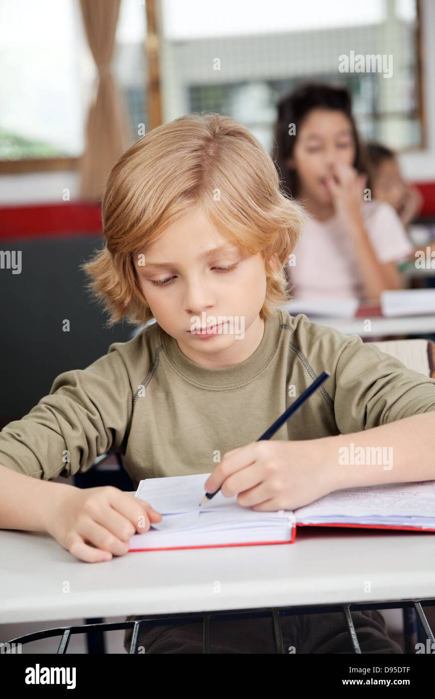 Cute little schoolboy writing in book at desk with classmates in ...