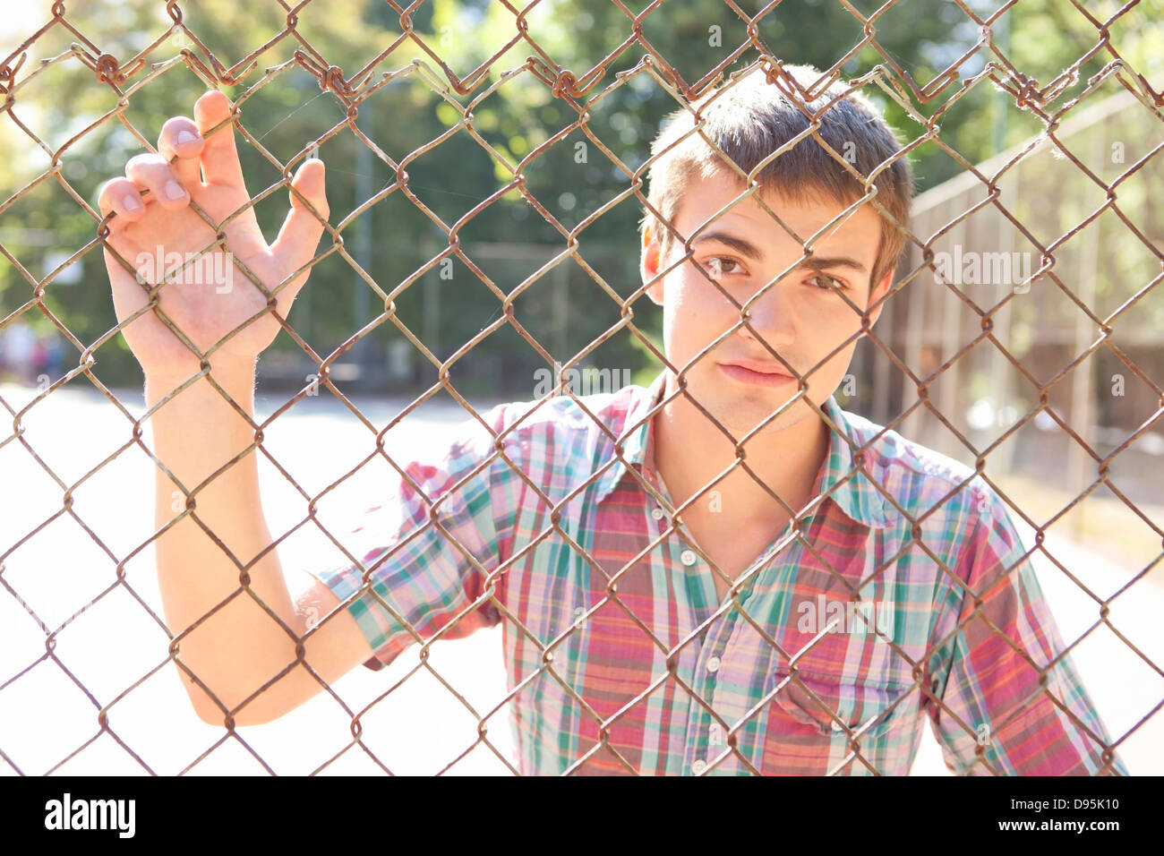 Portrait young man standing behind chain link fence in park near tennis ...