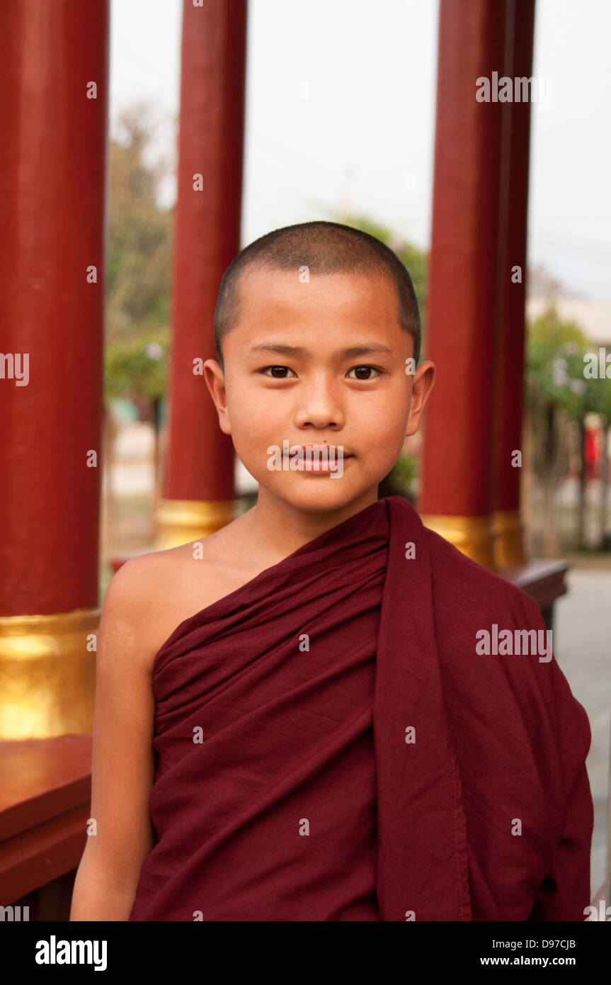 Boy monk in red robes stands in front of red temple pillars Myanmar ...