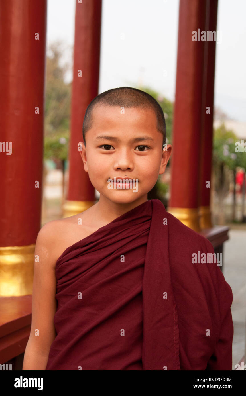 Boy monk in red robes stands in front of red temple pillars Myanmar ...