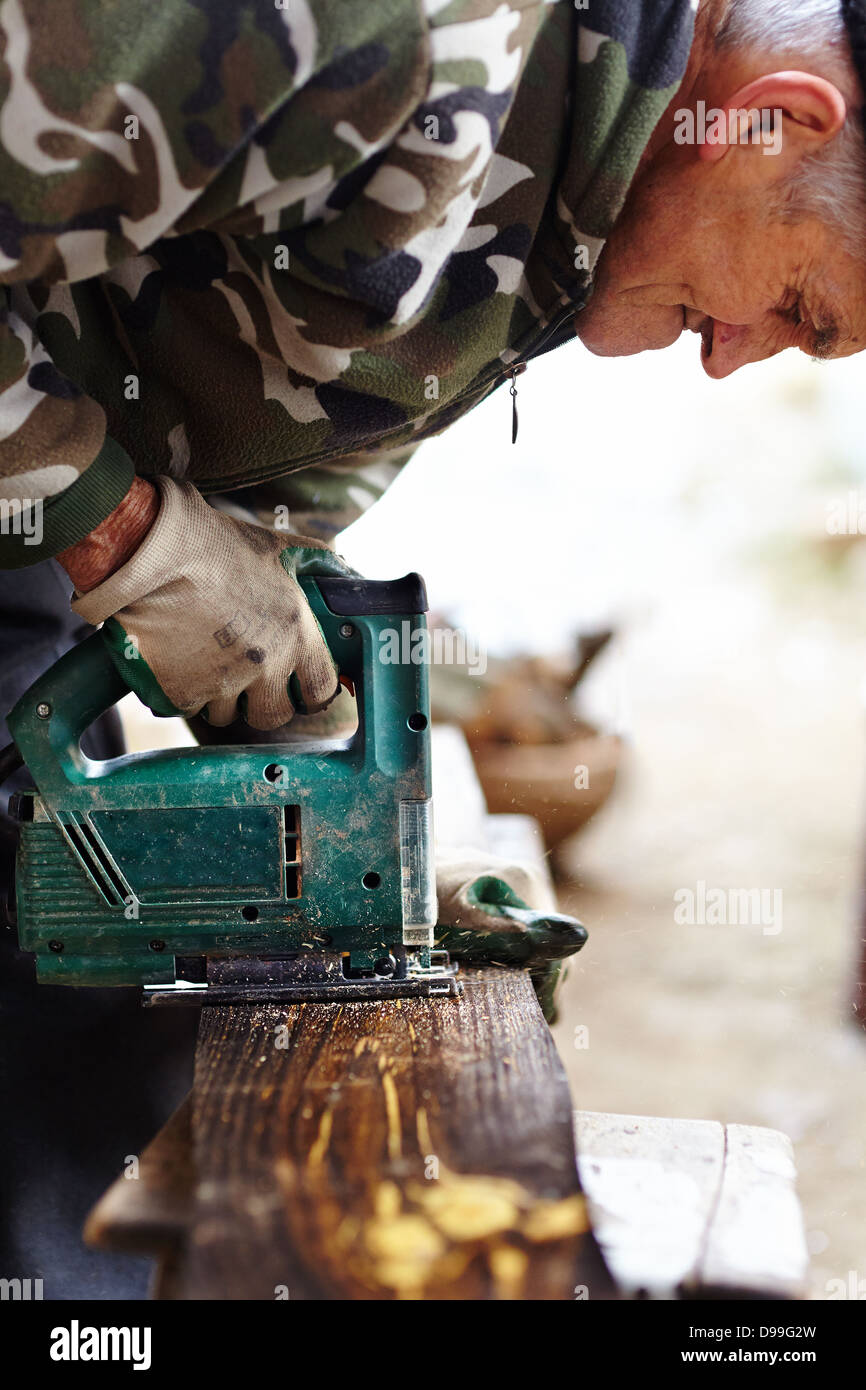 Old man with protection gloves using an electric saw to cut a plank ...