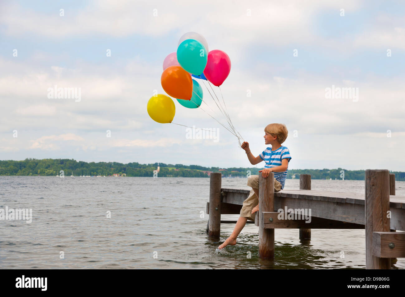 Germany, Bavaria, Boy sitting on pier and holding colourful balloons at ...
